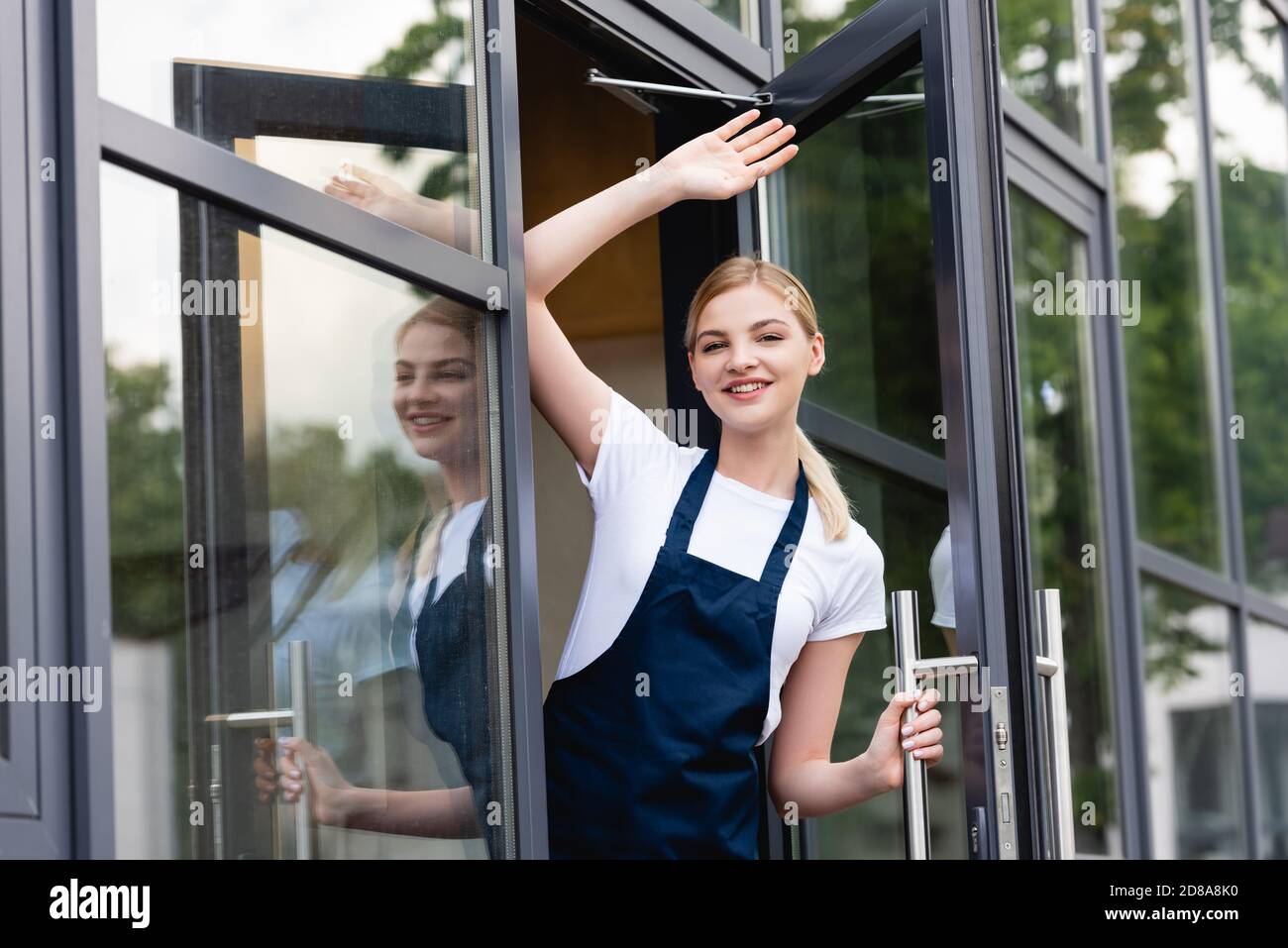 Smiling waitress waving hand at camera near open door of cafe Stock ...