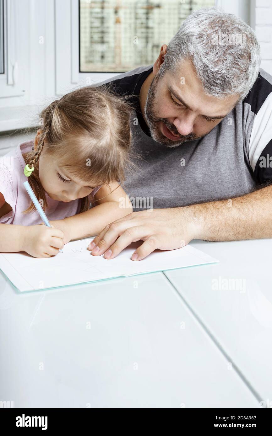 adult gray-haired father teaches little daughter to write at the table ...