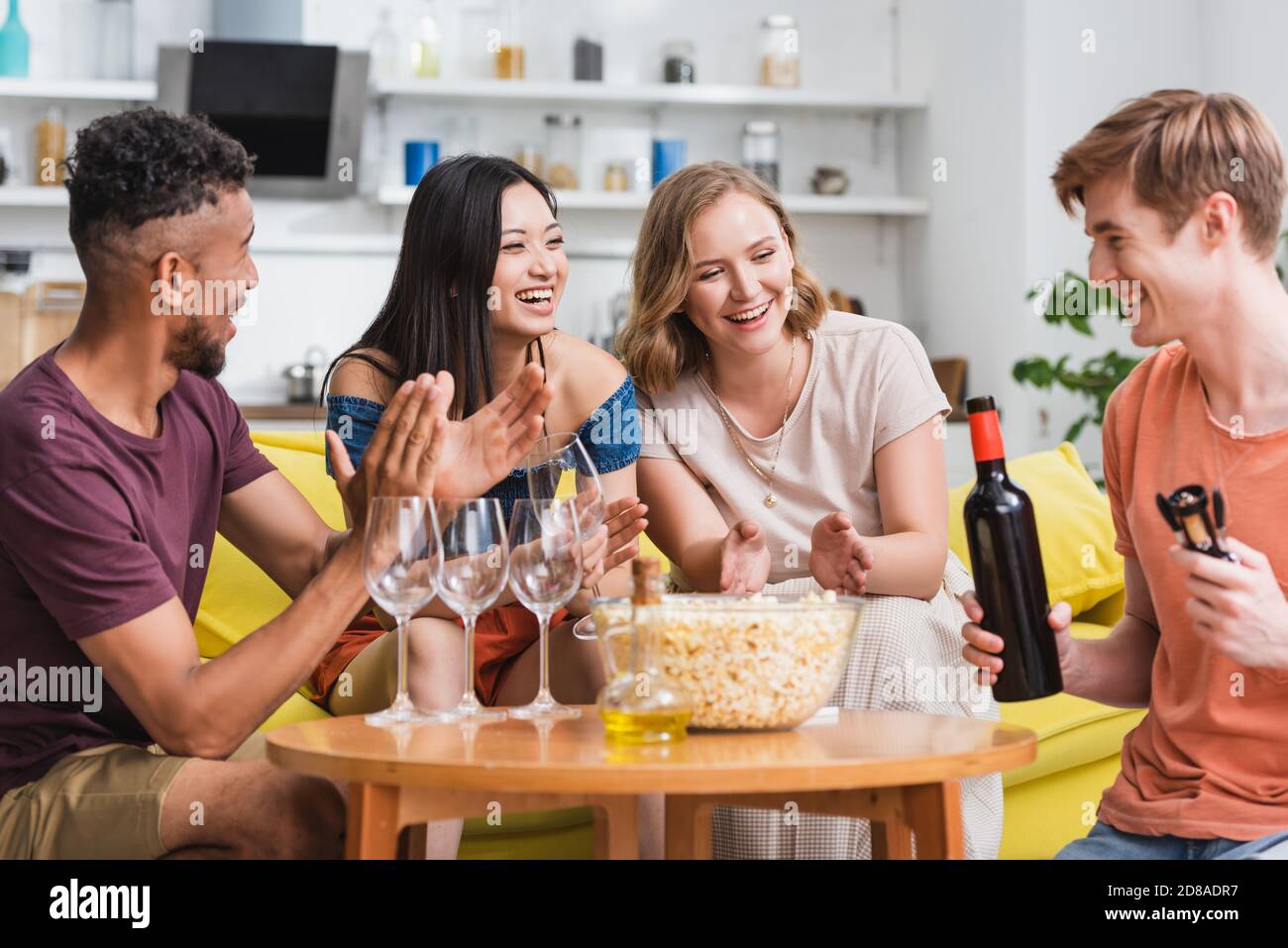 young man holding bottle of red wine near excited multicultural friends ...