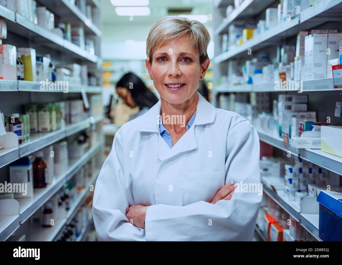 Smiling pharmacist with cross arms standing in medication isle wearing ...