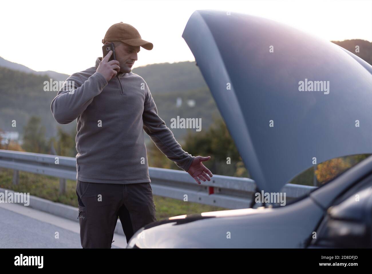 Angry young man waiting a help while sitting near the broken car at the ...