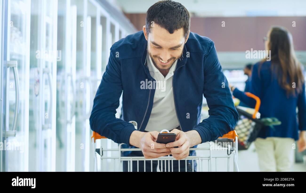 At the Supermarket: Handsome Man Uses Smartphone and Smiles, while ...
