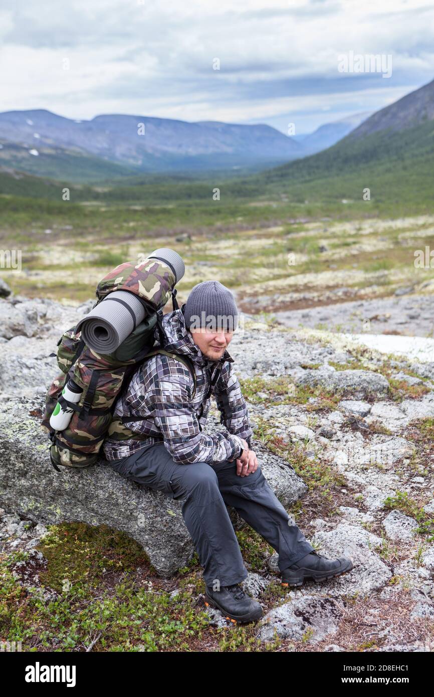 Adult Caucasian mountaineer resting on stone with heavy backpack on his ...