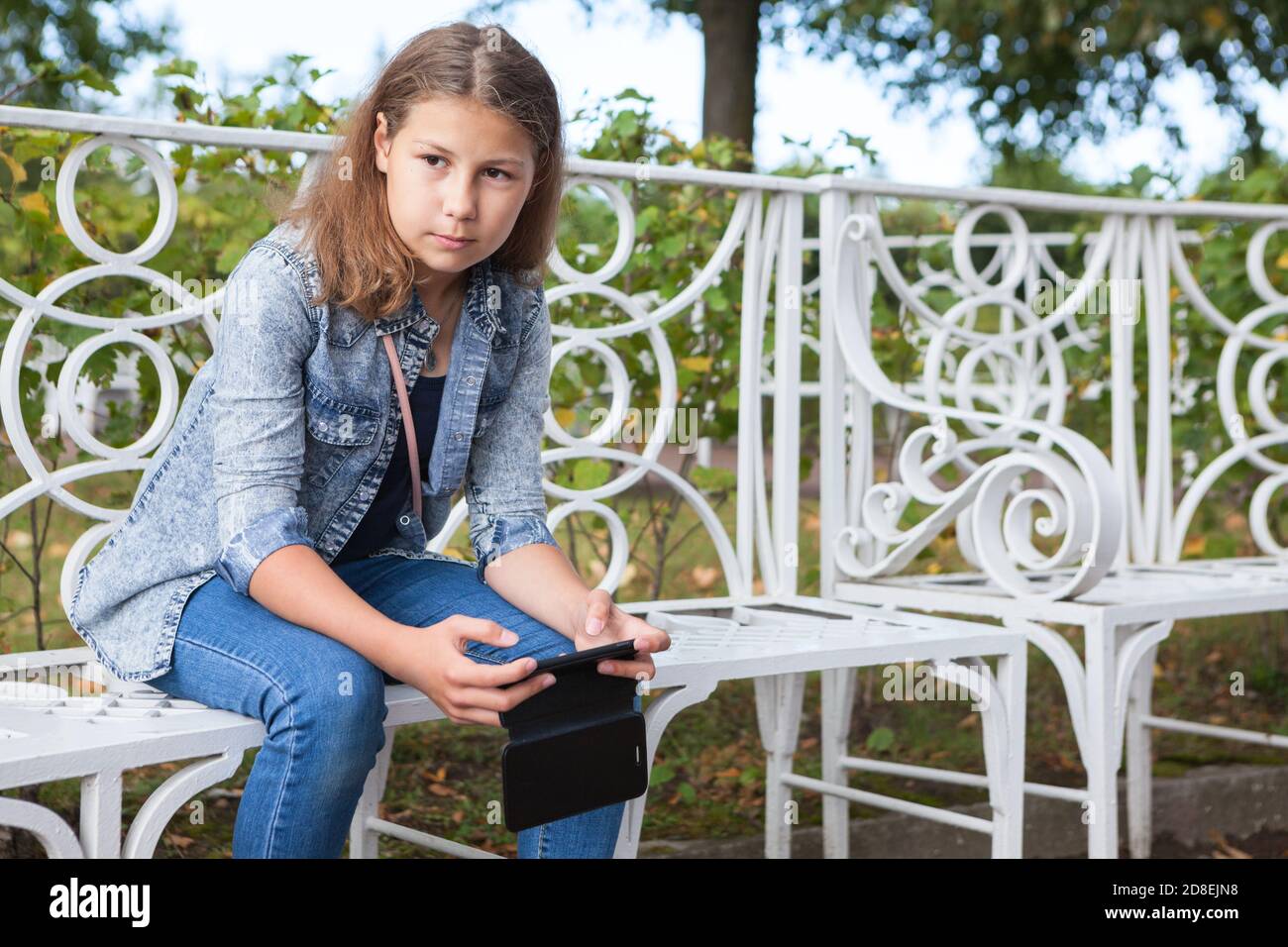 Thoughtful teen age girl sitting on bench with smartphone in hands ...