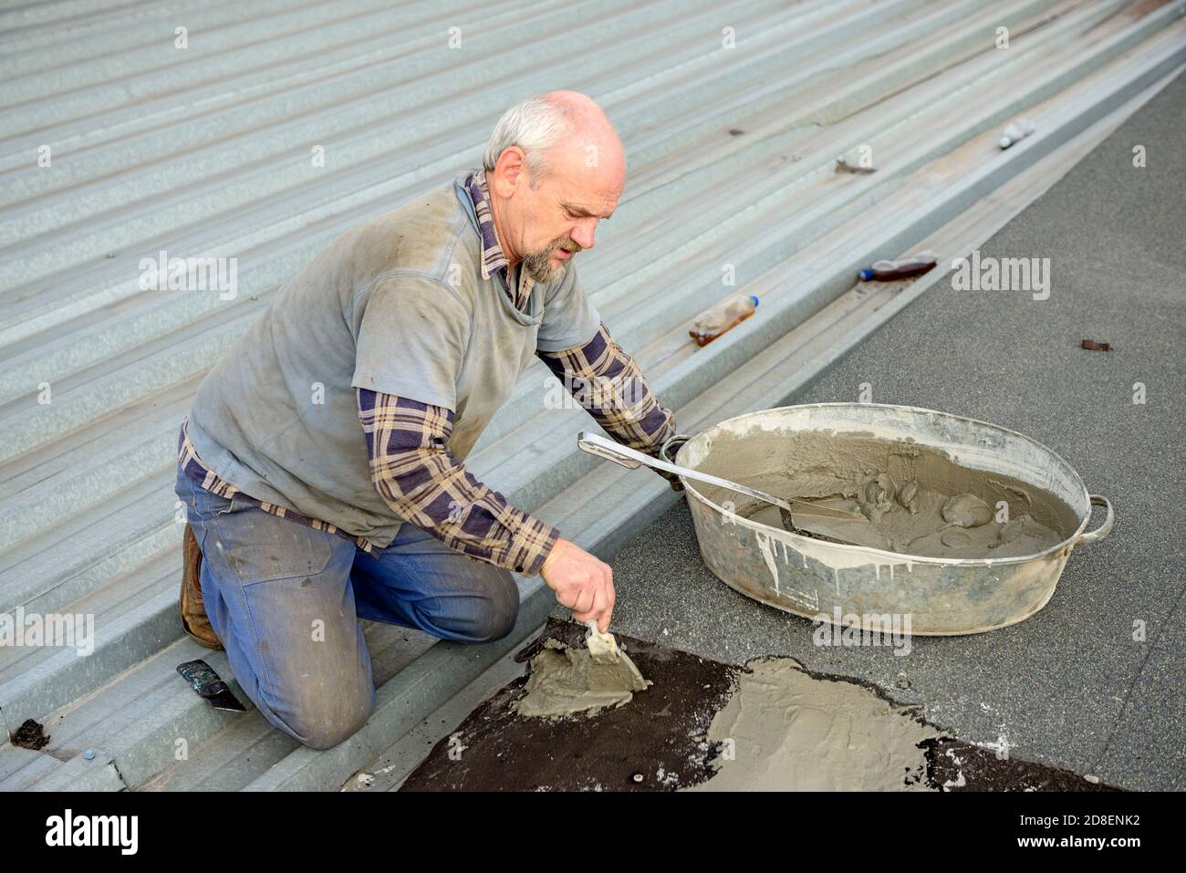 Kneeling aged home roofer is using up mortar in basin for repairing of ...