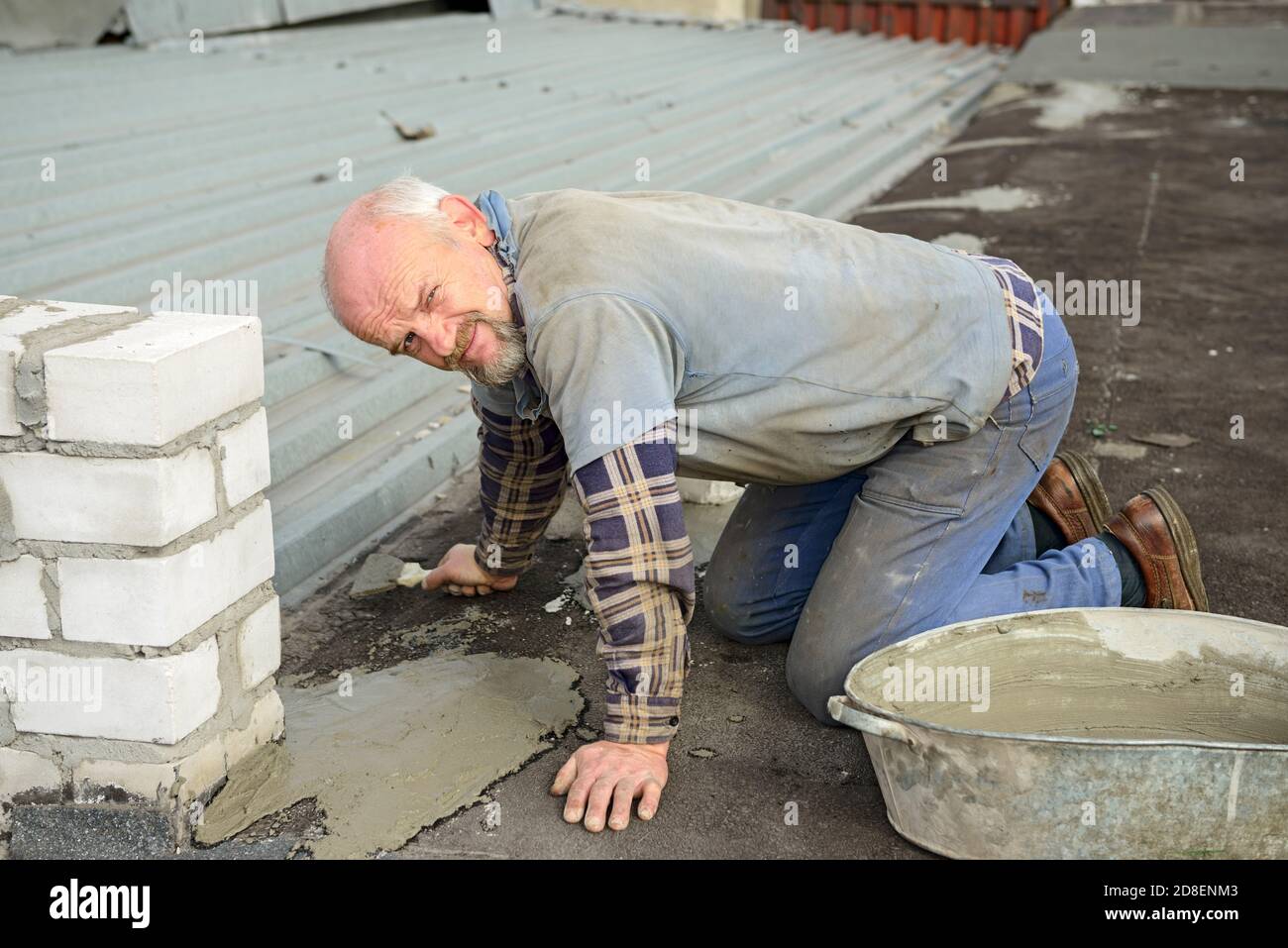 Aged home roofer is on all fours using up mortar in basin for repairing ...