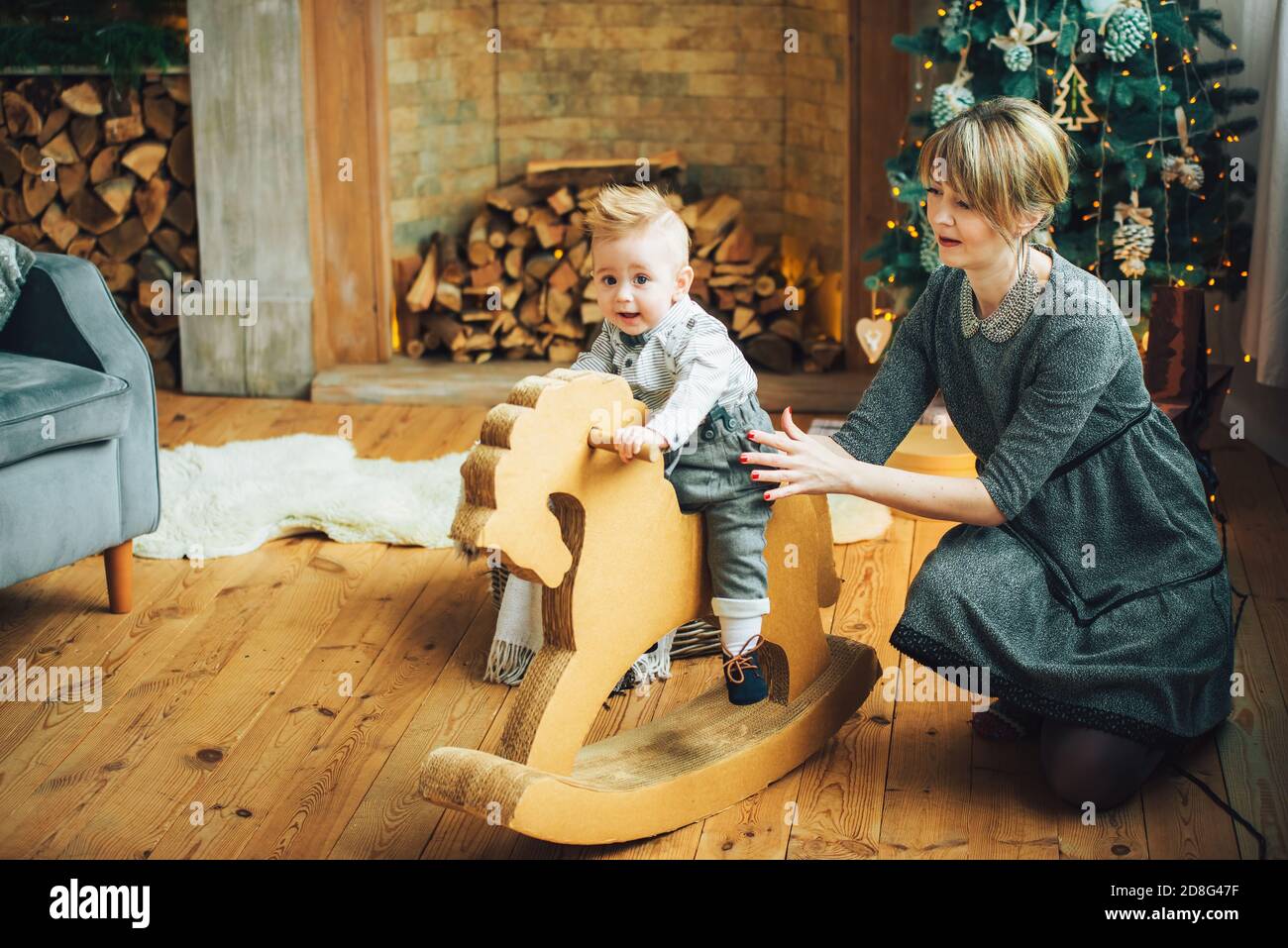 Small boy riding rocking horse in front of christmas tree. Mother ...