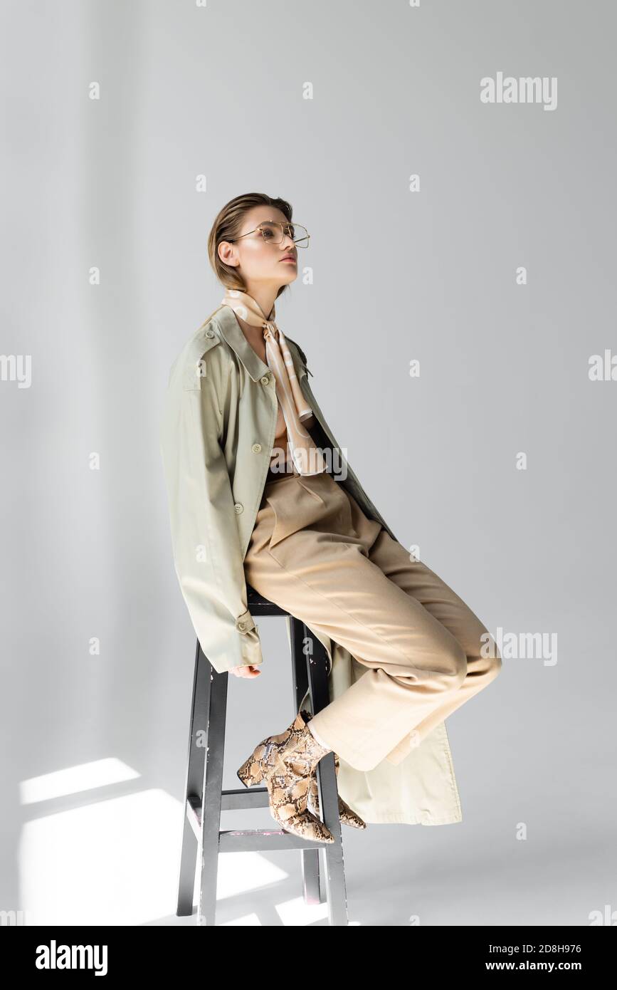 full length of young woman in glasses, trench coat and scarf sitting on ...