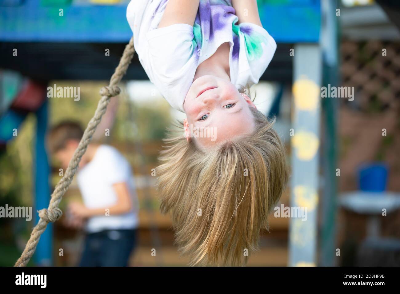 Beautiful little girl hangs upside down on the playground. Little girl smiles into the camera ...