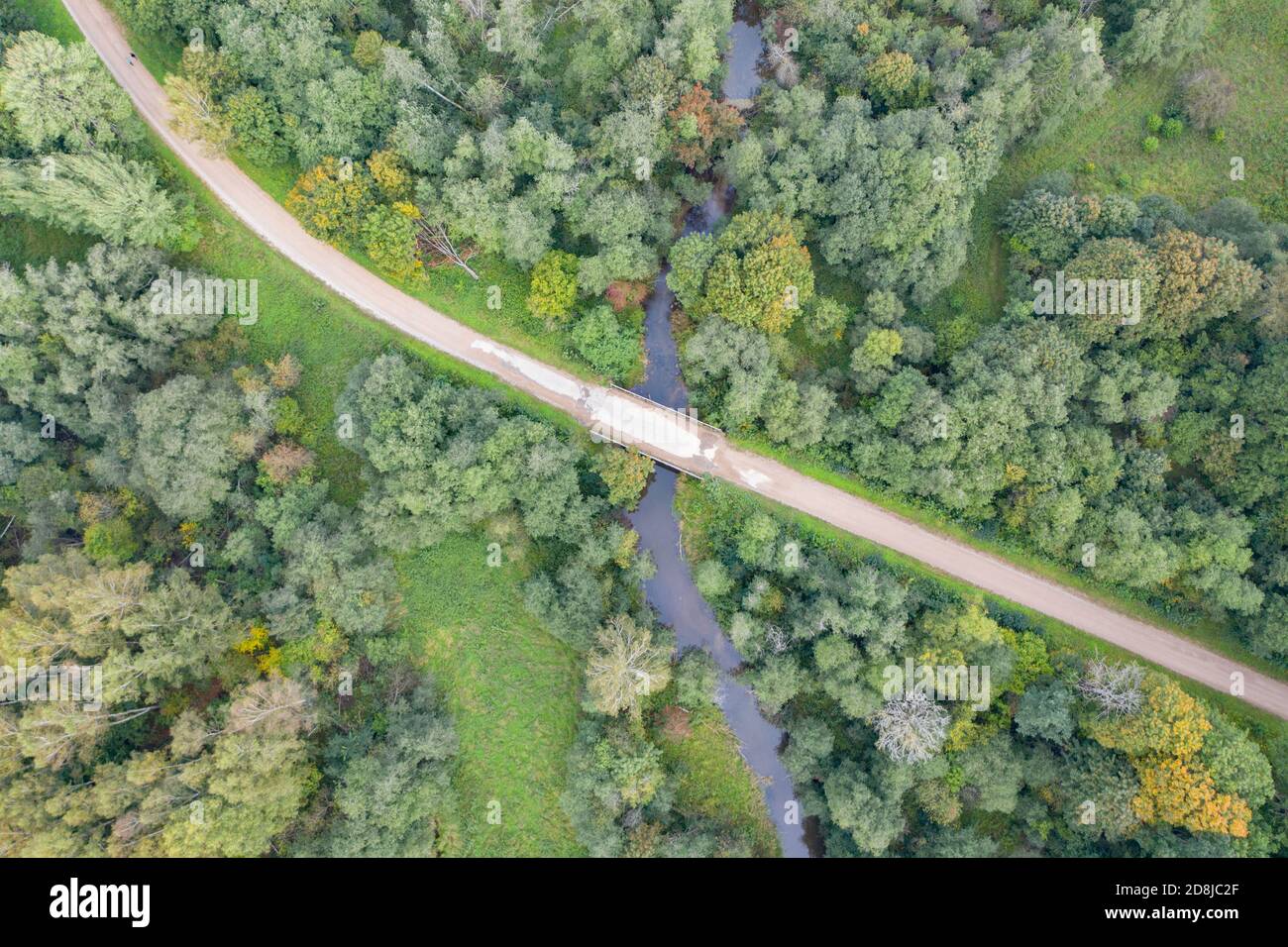 Aerial top down view of gravel road and bridge over winding river flowing through green forest ...