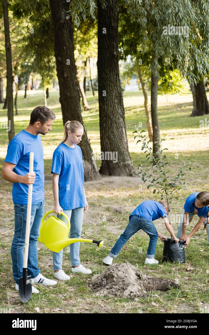 children carrying young tree while parents standing with shovel and ...