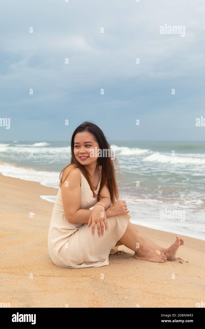 Happy Asian woman sitting on the sand near the water and enjoying the ...