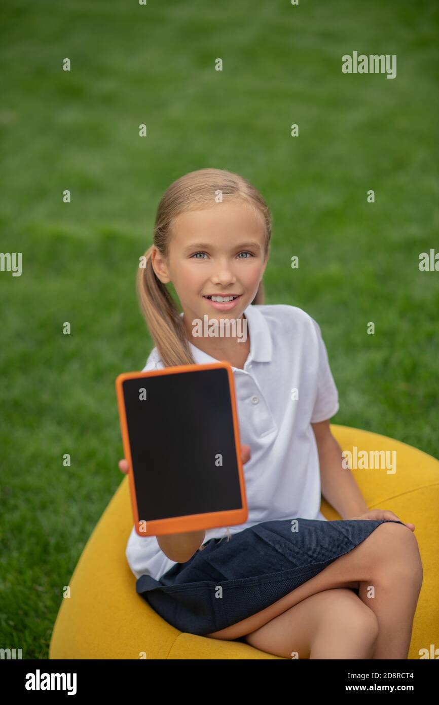 Cute fair-haired schoolgirl sitting on a bag chair and showing her tablet Stock Photo - Alamy