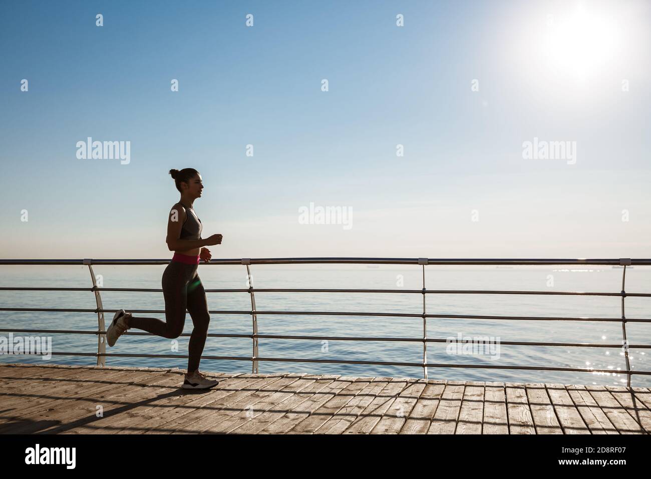 Side view of young fitness woman running along pier. Female athlete ...