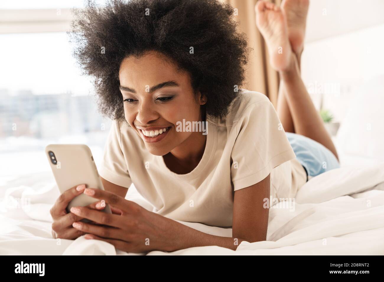 Young smiling african woman sending text messages, laying in bedroom ...