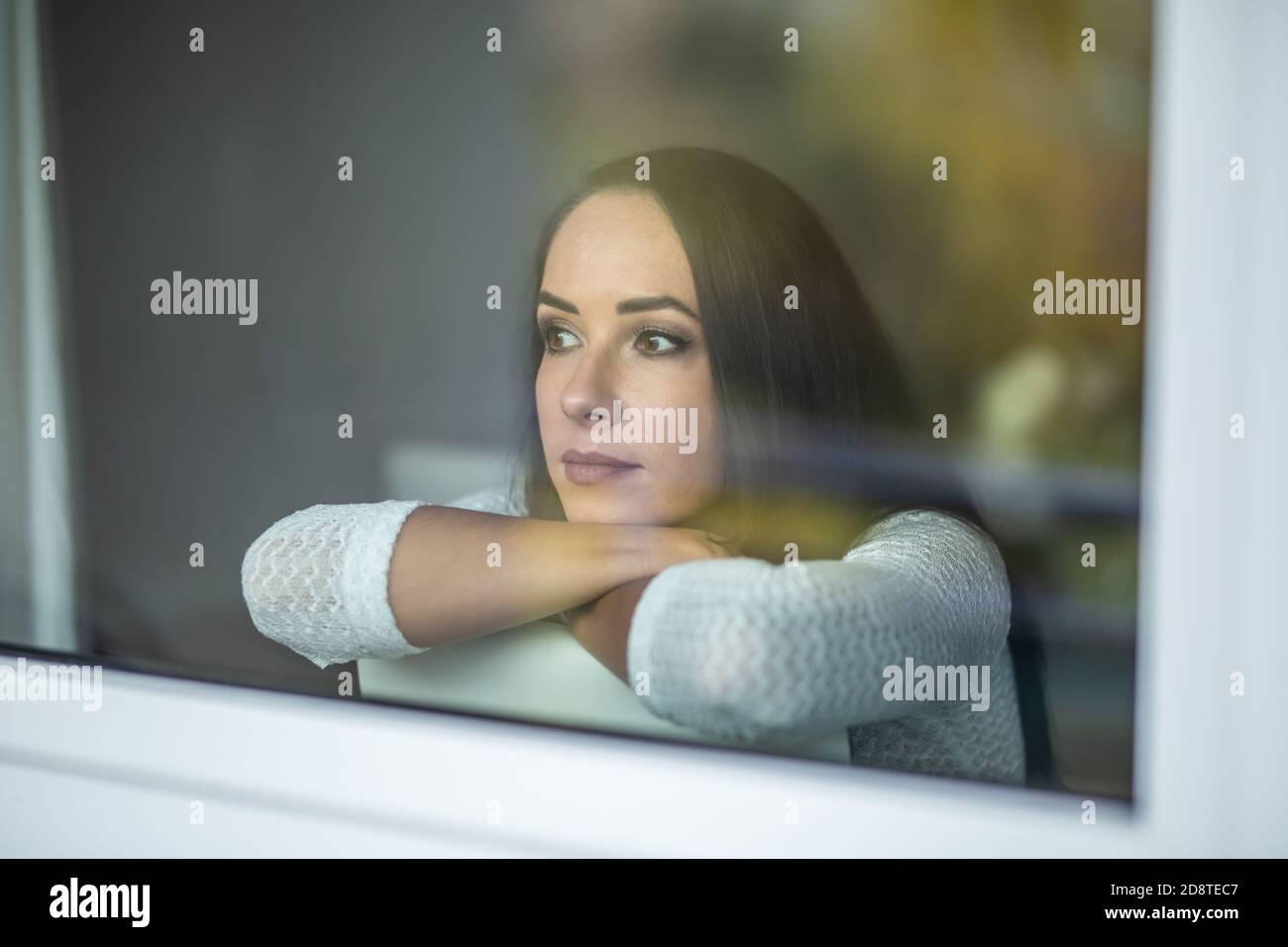 Sad good-looking female sits on a chair behind the window looking ...