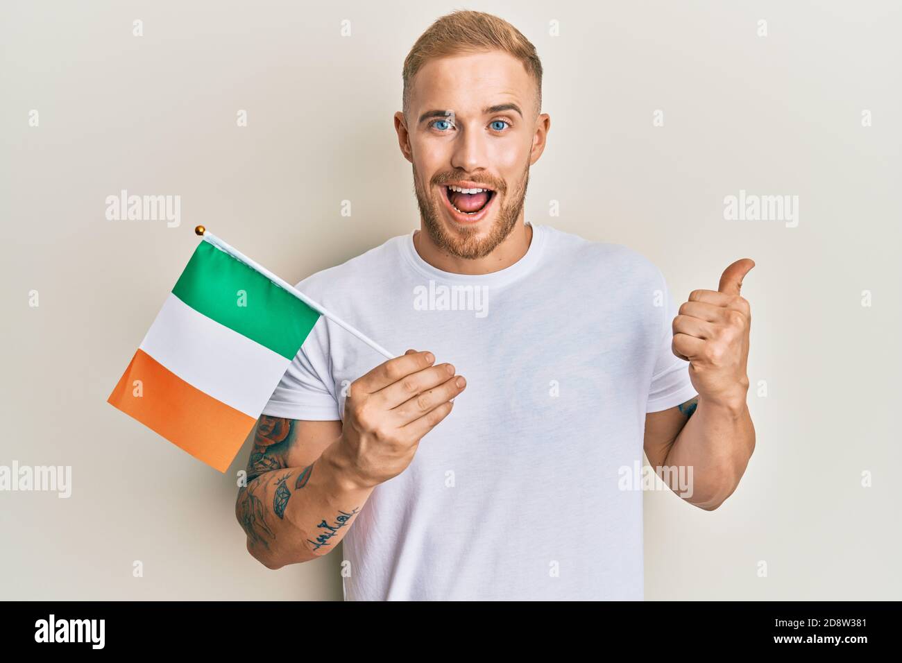 Young caucasian man holding irish flag pointing thumb up to the side ...