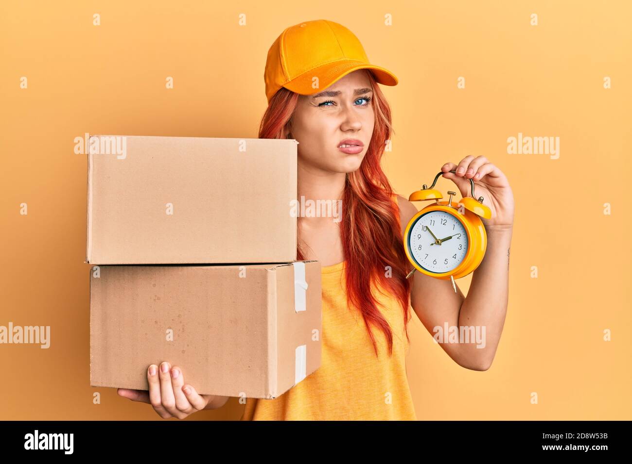 Young redhead woman holding delivery package and alarm clock clueless ...