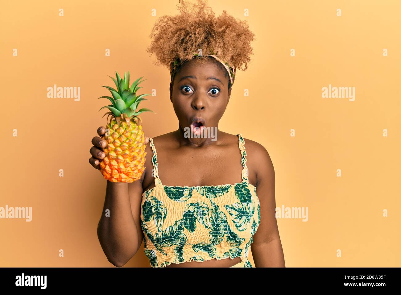 Young african woman with afro hair holding pineapple scared and amazed ...