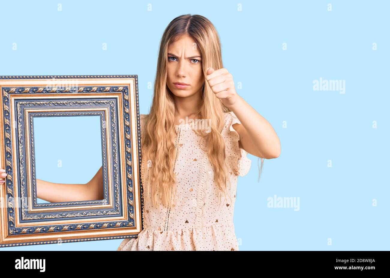 Beautiful caucasian woman with blonde hair holding empty frame annoyed ...