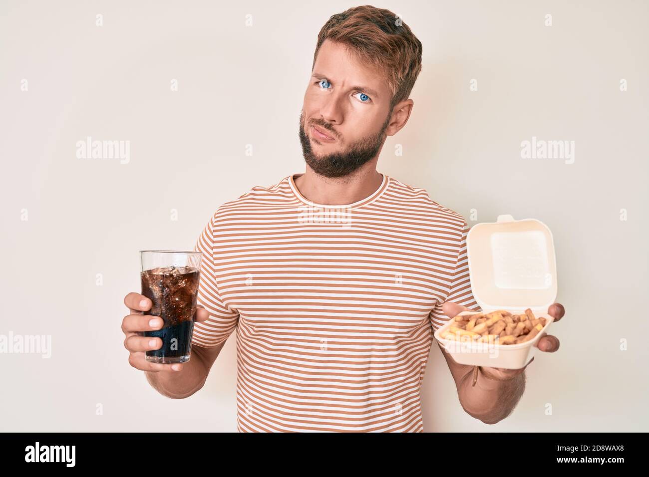 Young caucasian man eating fast food in shock face, looking skeptical ...