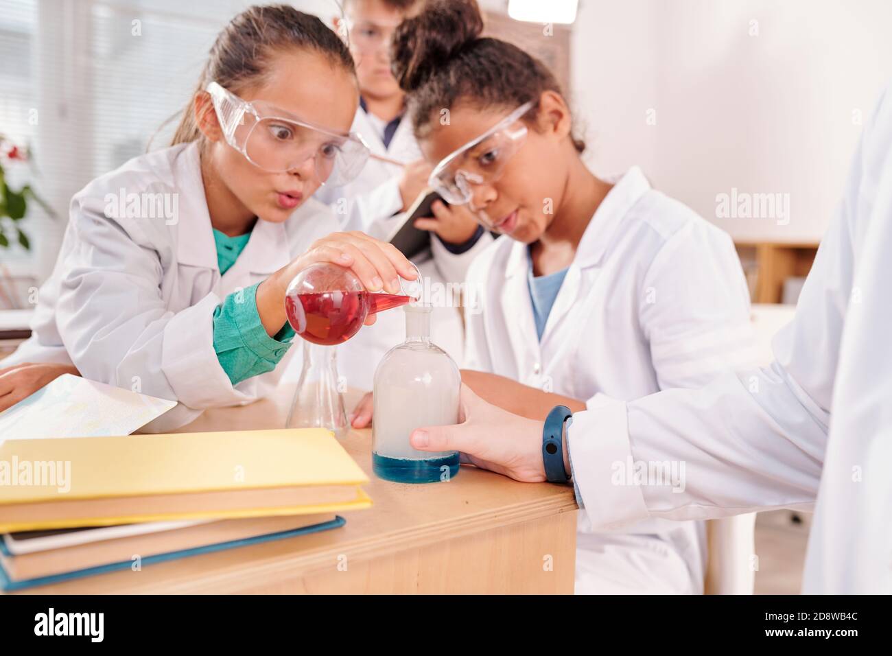 Amazed schoolgirls in protective eyeglasses and whitecoats looking at ...