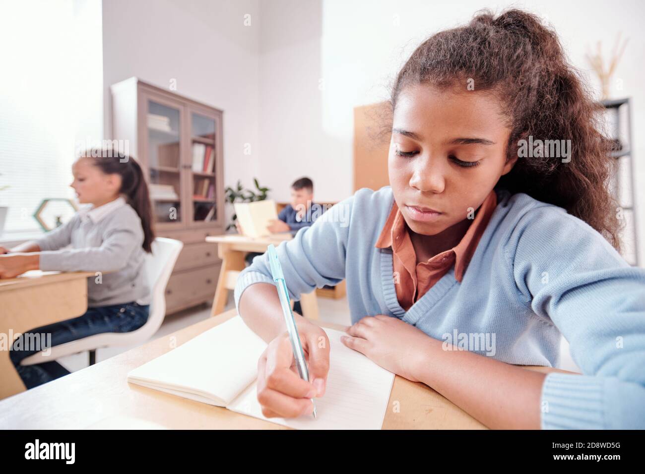 Serious schoolgirl making notes or writing essay while sitting by desk ...