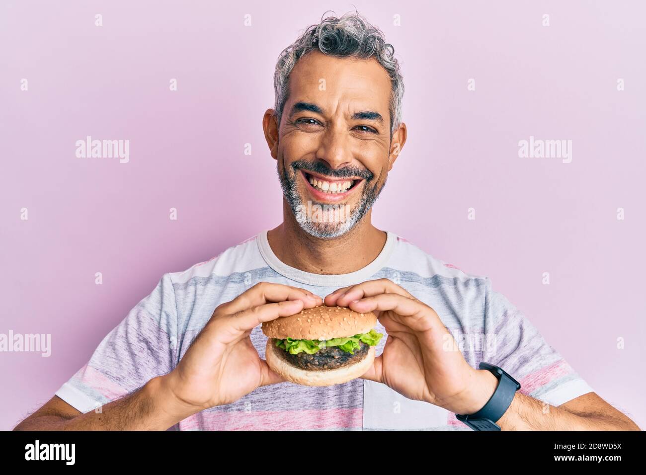Middle age grey-haired man eating a tasty classic burger smiling with a ...