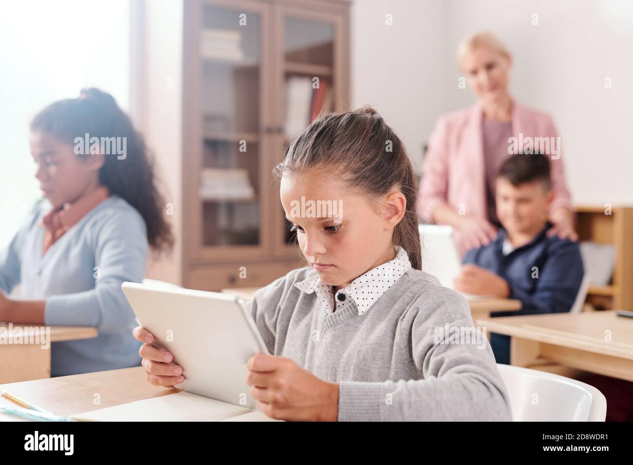 Cute girl and her classmates with tablets working individually with ...