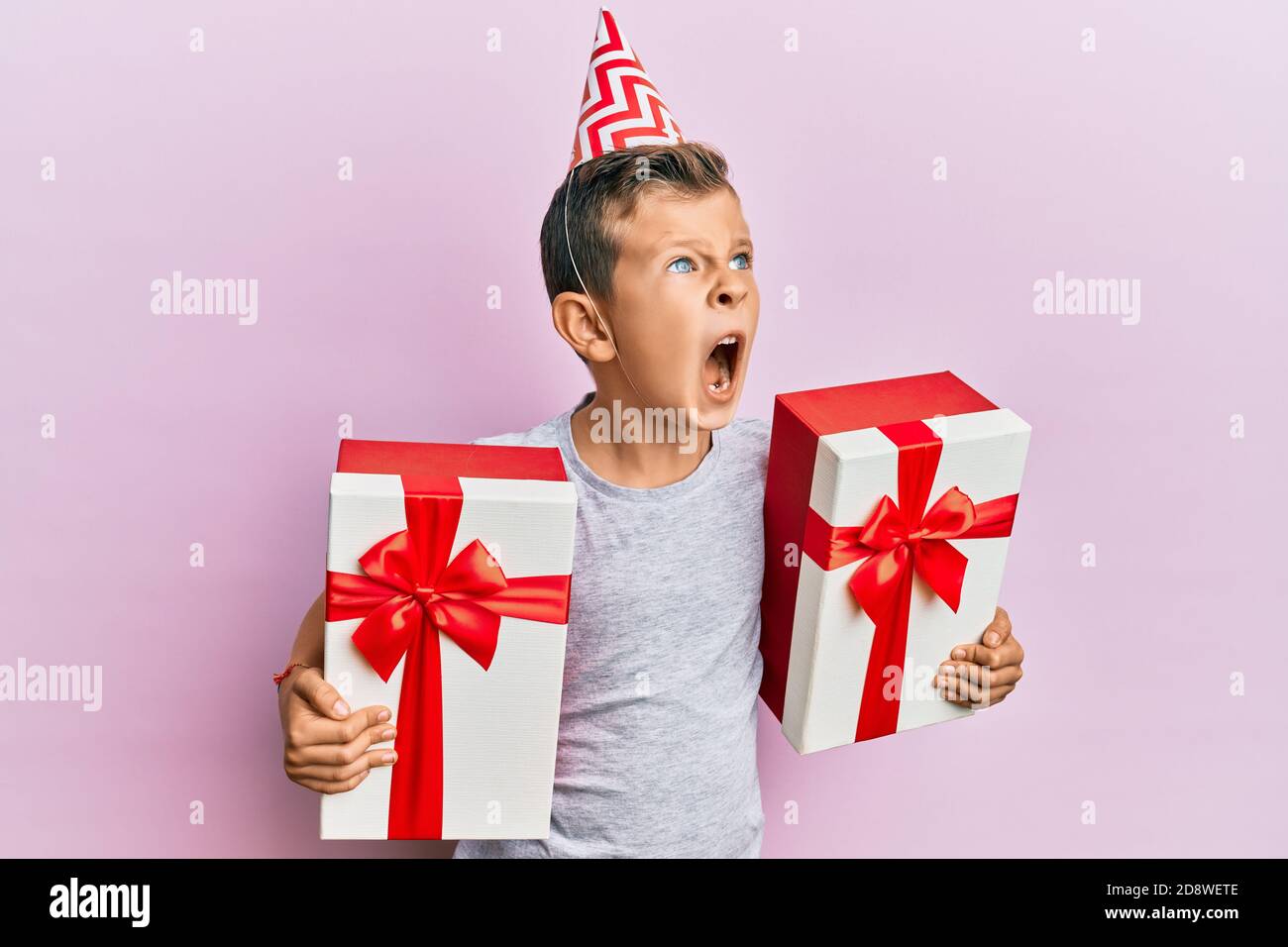 Adorable caucasian kid wearing birthday hat holding presents angry and ...