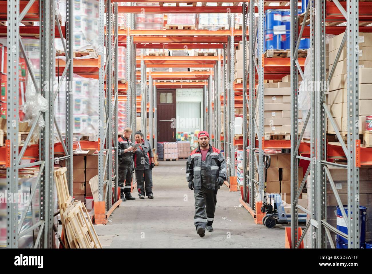 Young mixed-race warehouse worker in uniform moving along aisle between ...