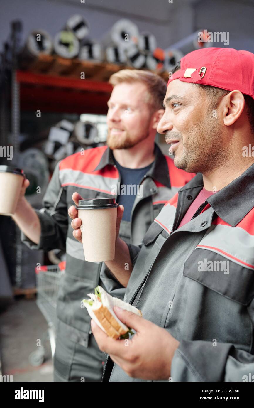 Two happy young warehouse workers having sandwiches and coffee at lunch ...