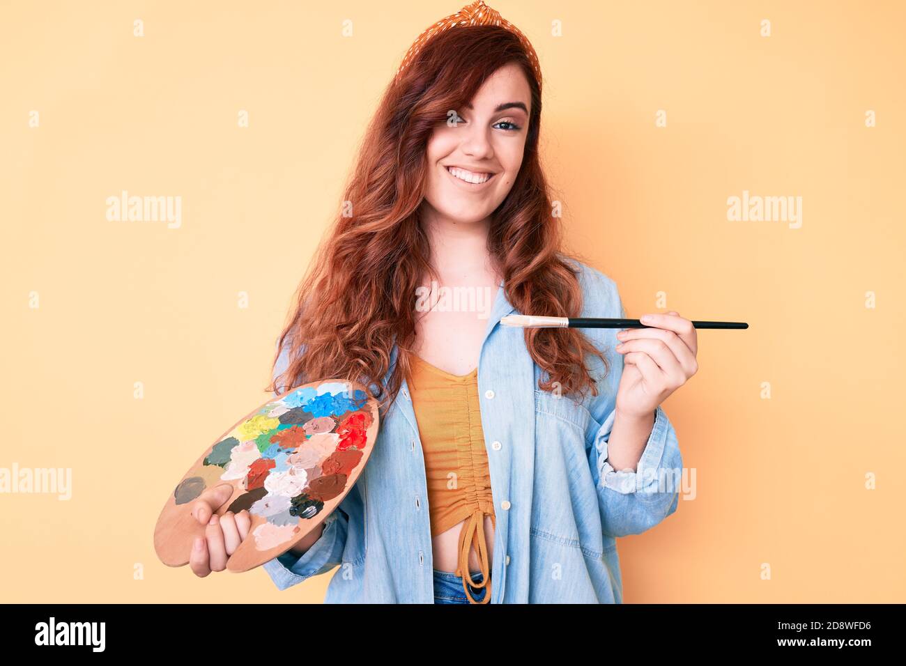 Young beautiful woman holding paintbrush and palette looking positive ...