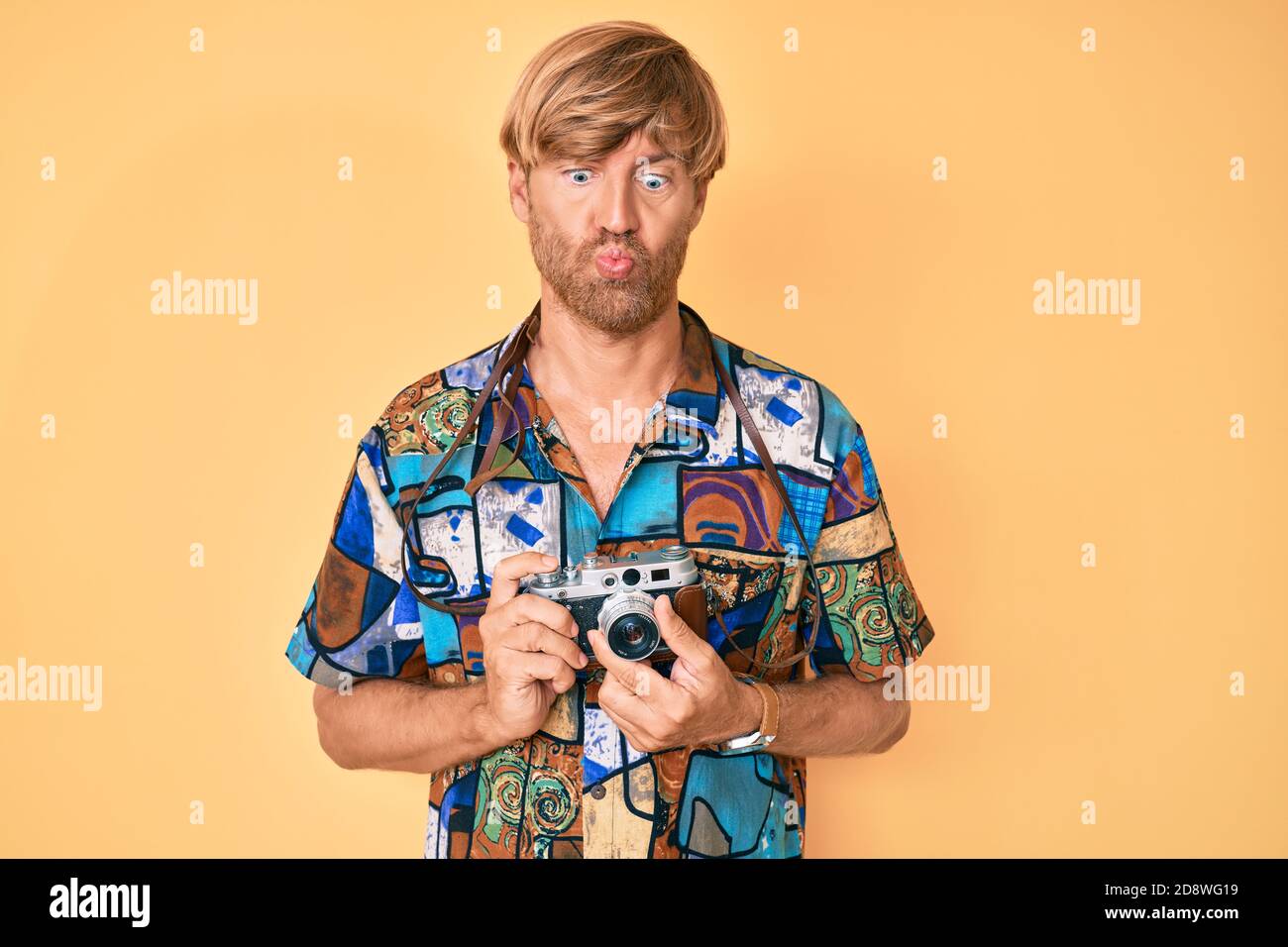 Young blond man holding vintage camera making fish face with mouth and ...