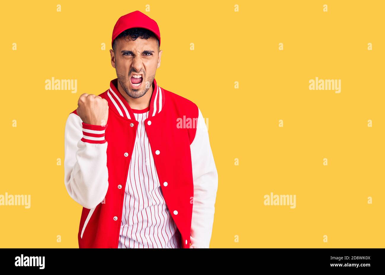 Young hispanic man wearing baseball uniform angry and mad raising fist ...