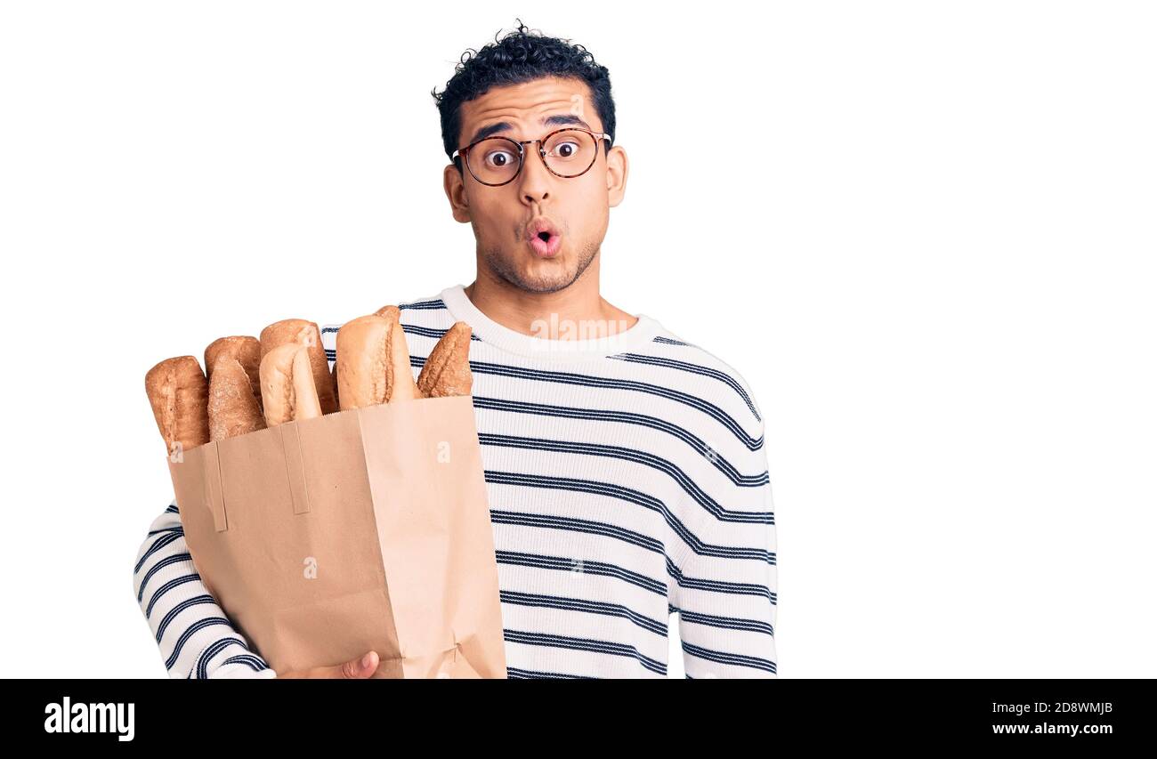 Hispanic handsome young man holding paper bag with bread scared and ...