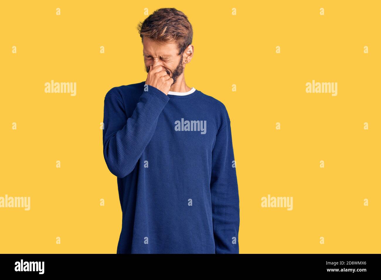 Handsome blond man with beard wearing casual sweater smelling something ...