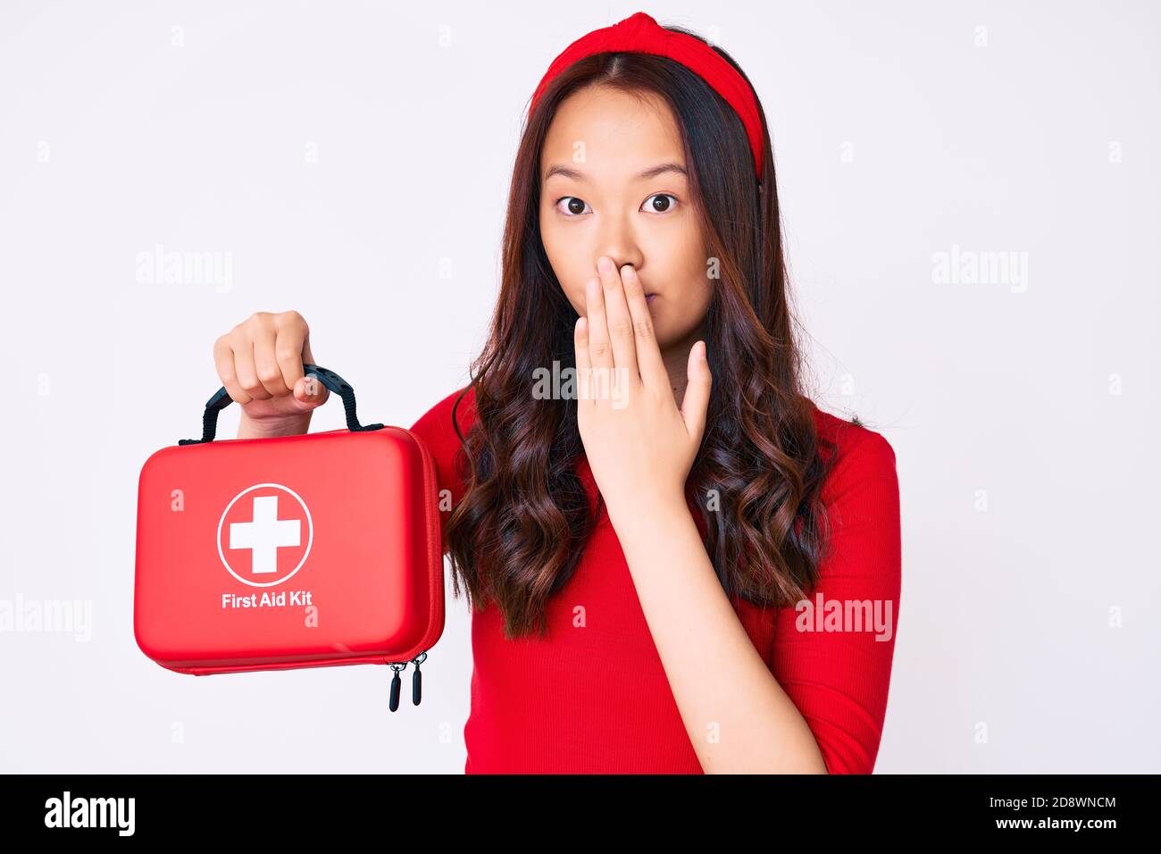 Young beautiful chinese girl holding first aid kit covering mouth with ...