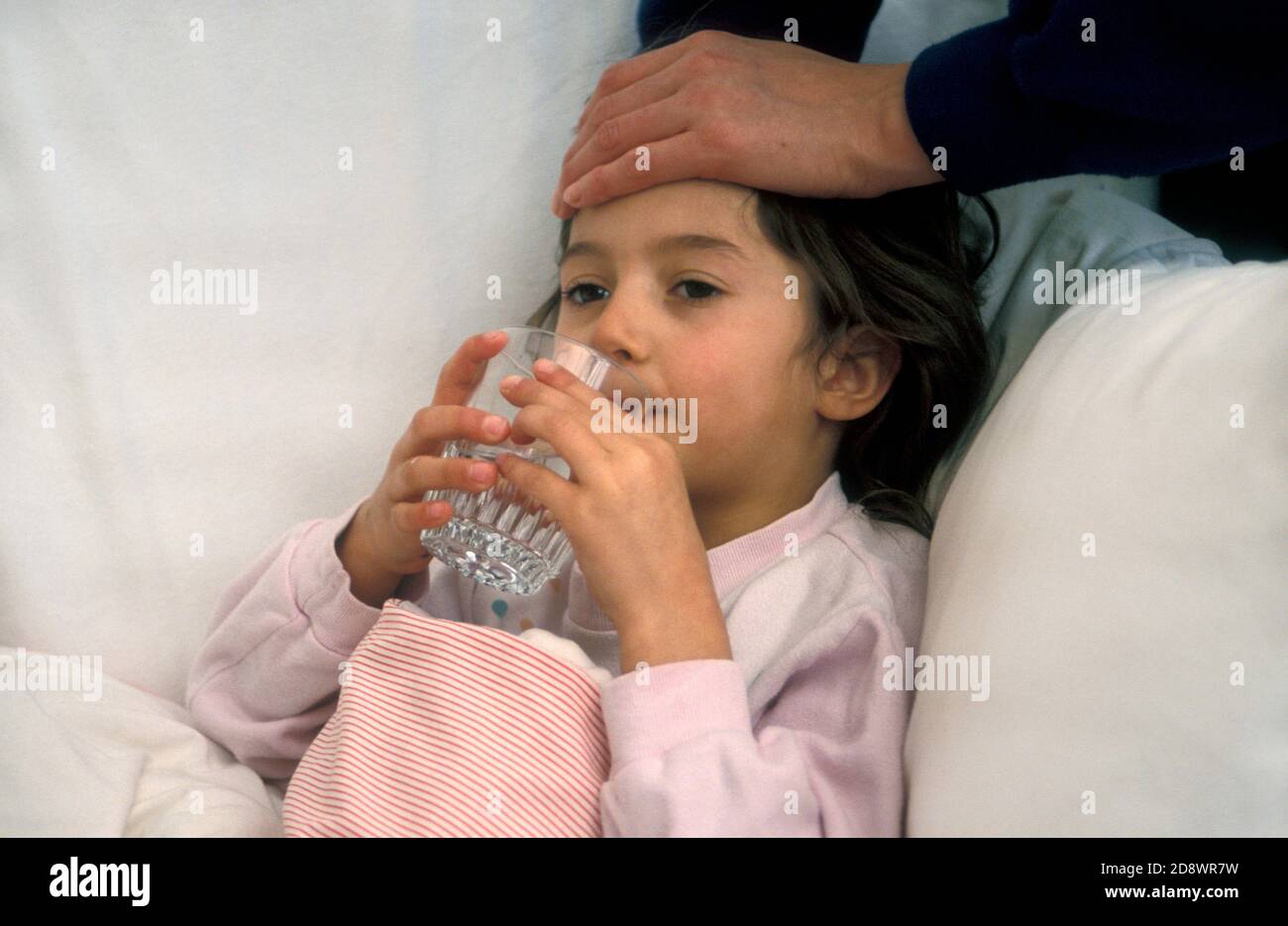 parents hand on the forehead of young girl in bed with a fever drinking ...