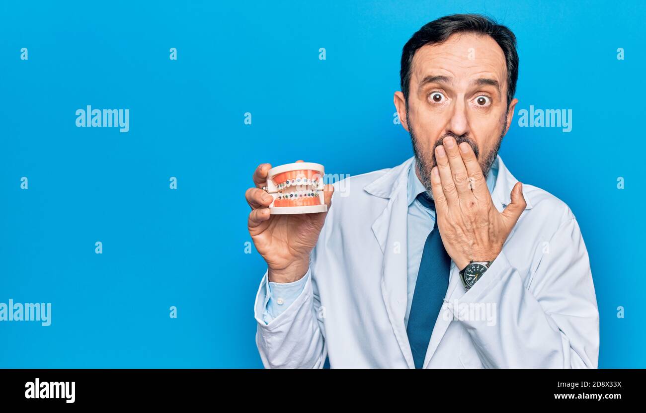 Middle age handsome dentist man wearing coat holding plastic teeth with ...