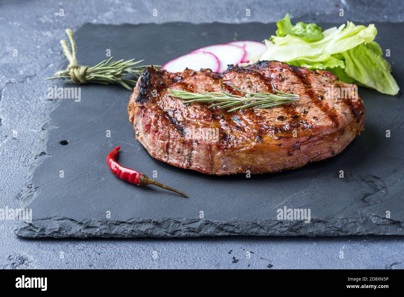 A ribeye steak with sprig of rosemary and radish on a black stone slab ...