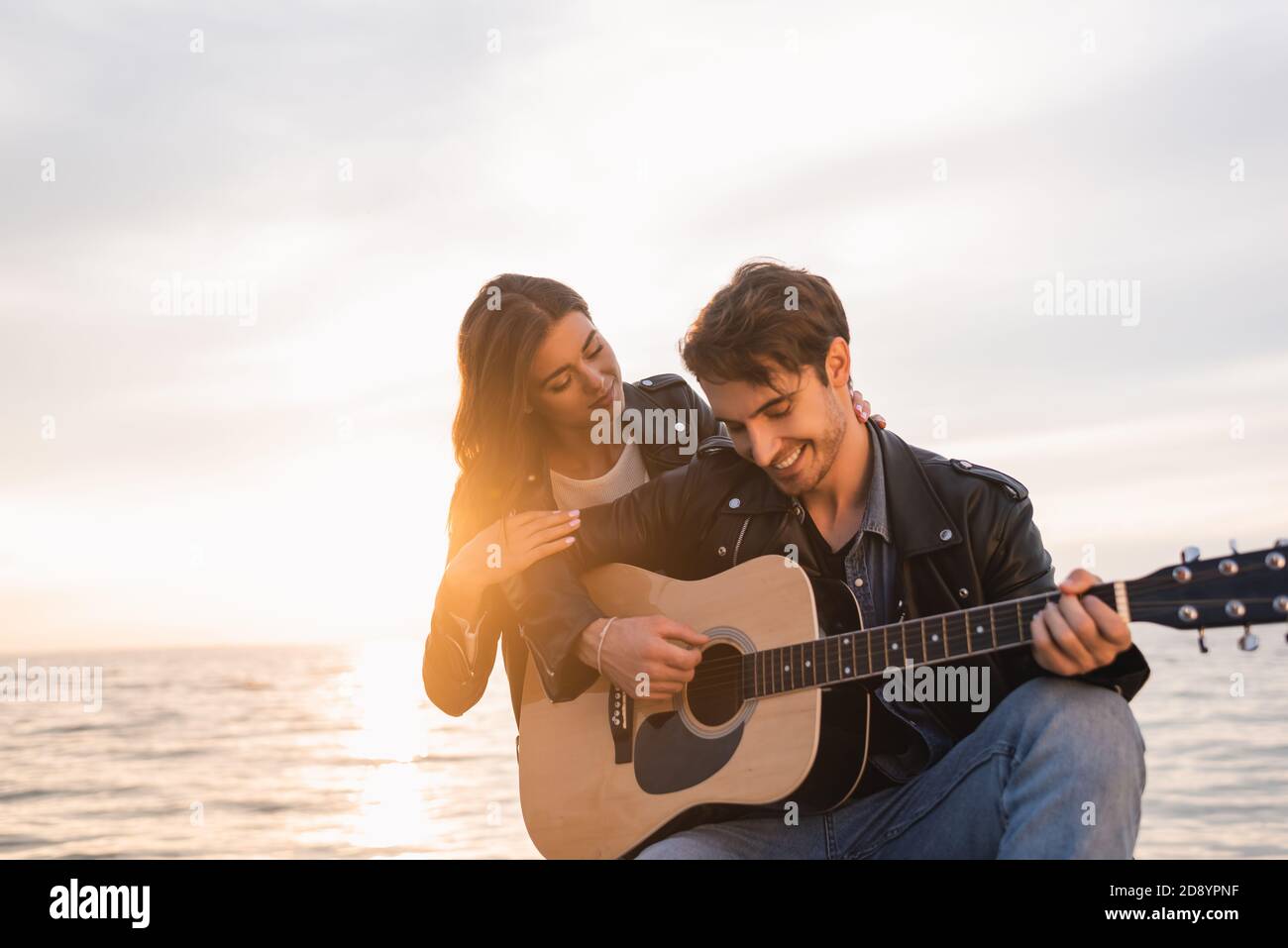 Woman hugging boyfriend playing acoustic guitar near sea and sunset at ...