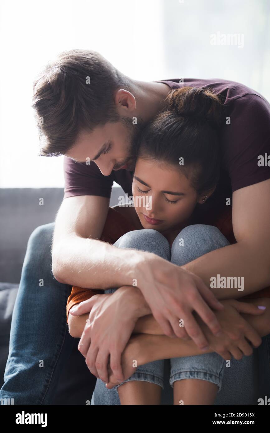young man embracing and calming beloved woman sitting on sofa with ...
