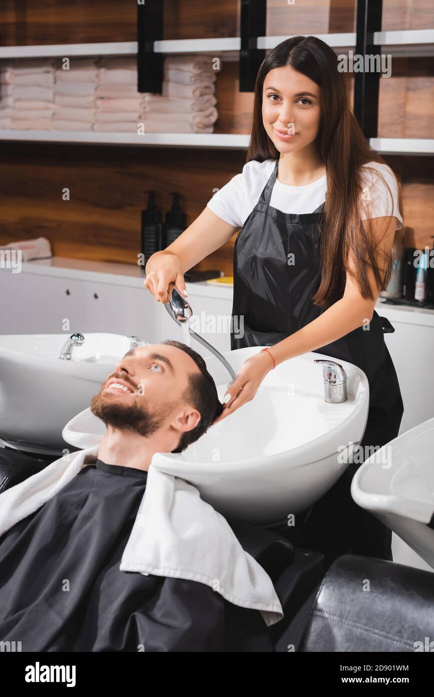 young barber in apron looking at camera while washing hair of happy ...