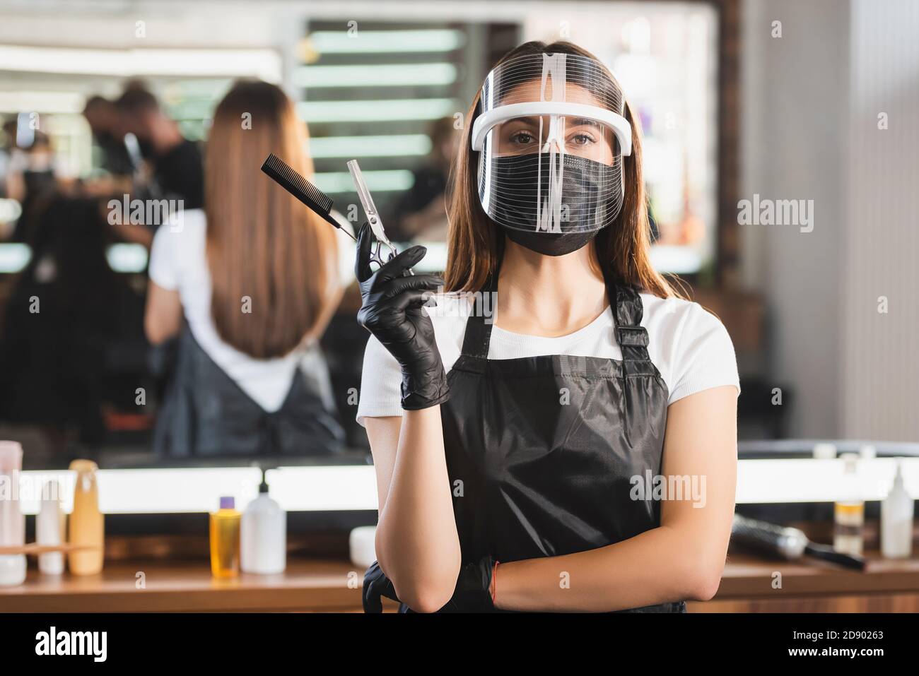 hairdresser in face shield, latex gloves and apron holding comb and ...
