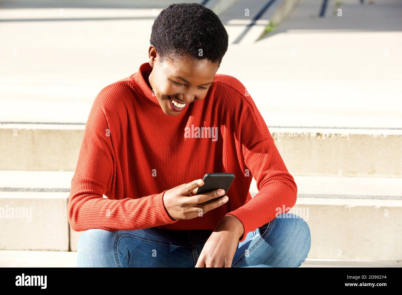 Portrait of smiling young black woman looking at cellphone outdoors ...