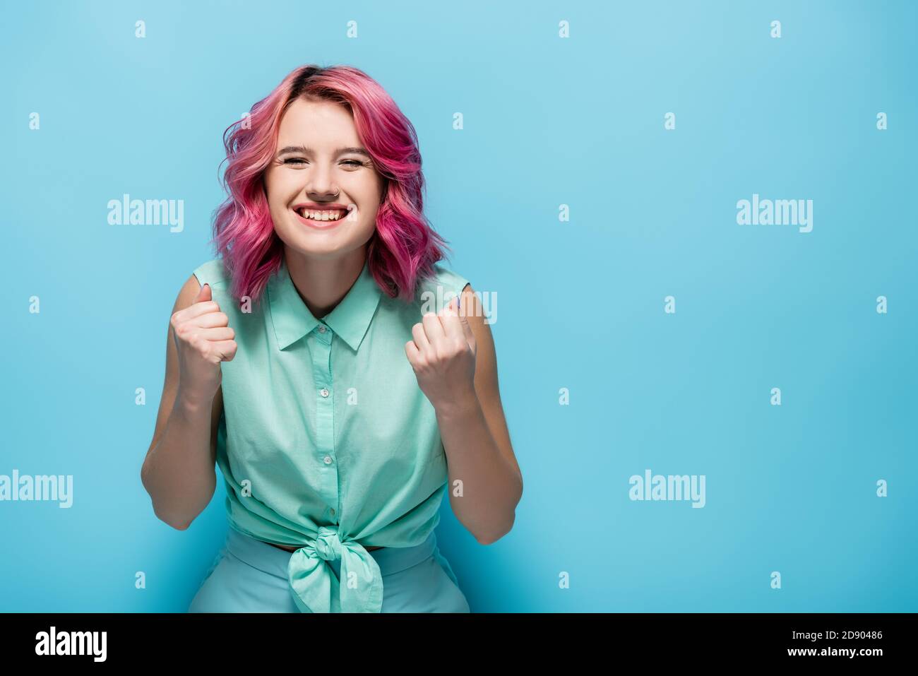 young woman with pink hair showing yeah gesture on blue background ...