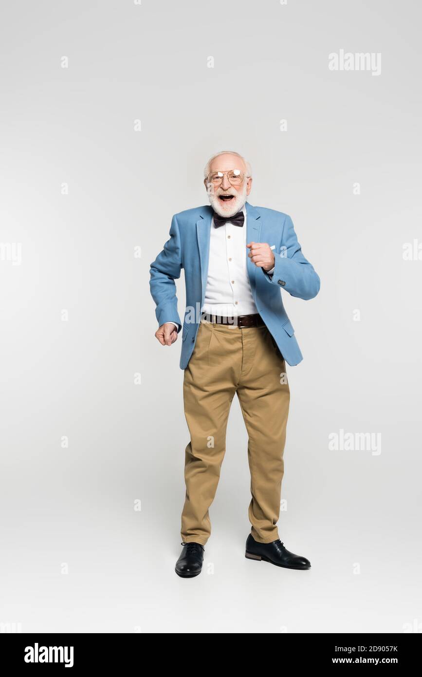 Cheerful senior man in jacket and bow tie dancing on grey background ...