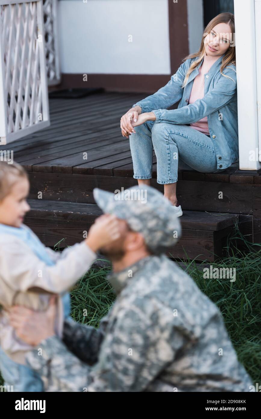 Smiling woman sitting on house threshold with blurred military ...