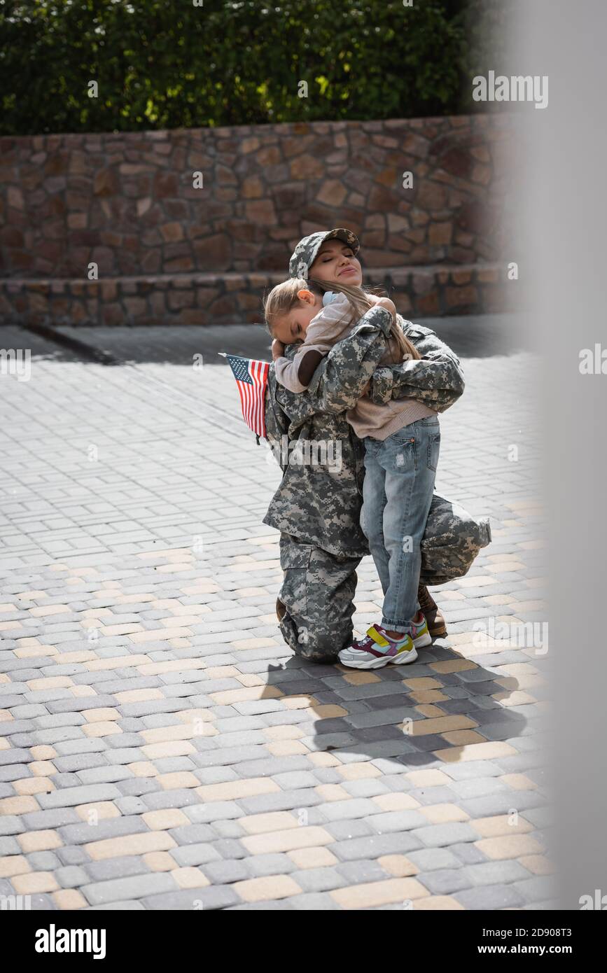 Smiling mother in military uniform sitting on knee and embracing ...